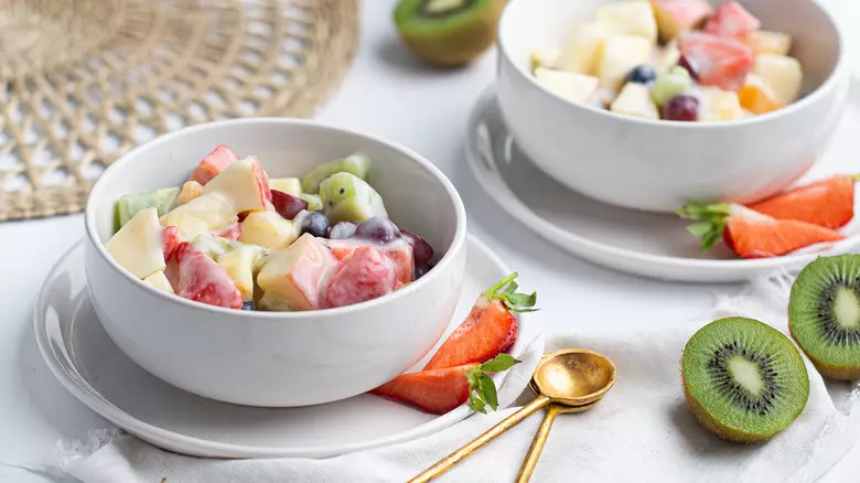 Dalawang puting mangkok ng fruit salad sa dalawang maliliit na puting plato sa tabi ng mga strawberry, kiwi, at gintong kutsara