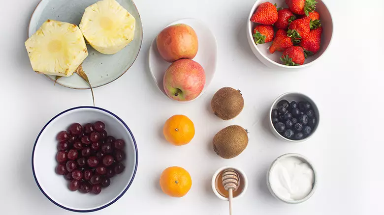 Gupitin ang mga pinya sa tabi ng mga pulang ubas, mansanas, tangerines, kiwi, pulot, yogurt, blueberries, at strawberry