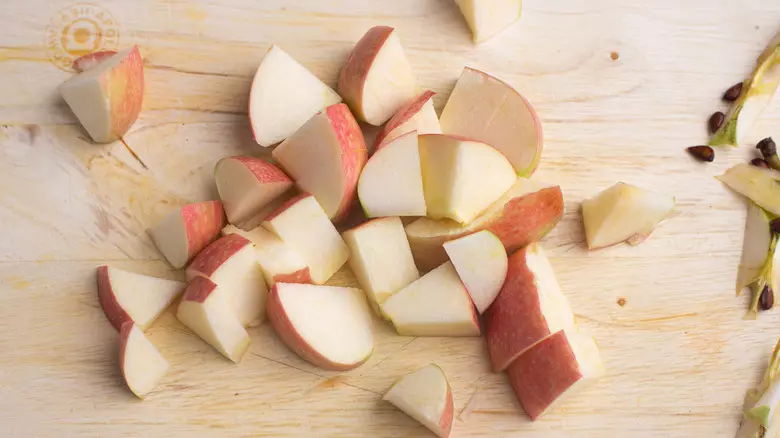 Isang kamay na may hawak na kutsilyo sa ibabaw ng pulang mansanas sa isang kahoy na cutting board