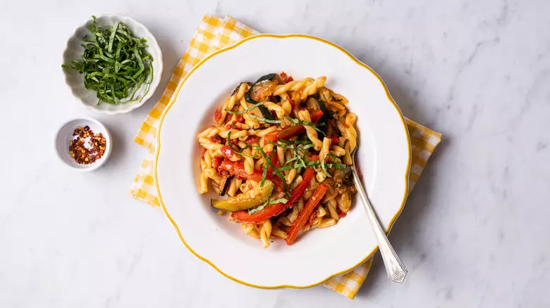 Summer vegetable pasta sa isang puting mangkok sa tabi ng basil at chili flakes.