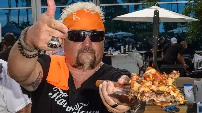 Guy Fieri me fashë portokalli, duke mbajtur një pjatë me BBQ