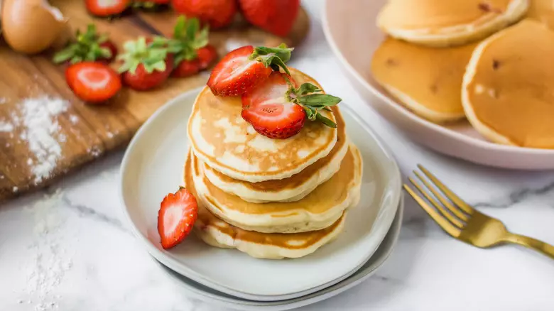 Un plato de panqueques de fresa con fresas junto a una tabla para cortar con fresas y un plato con más panqueques