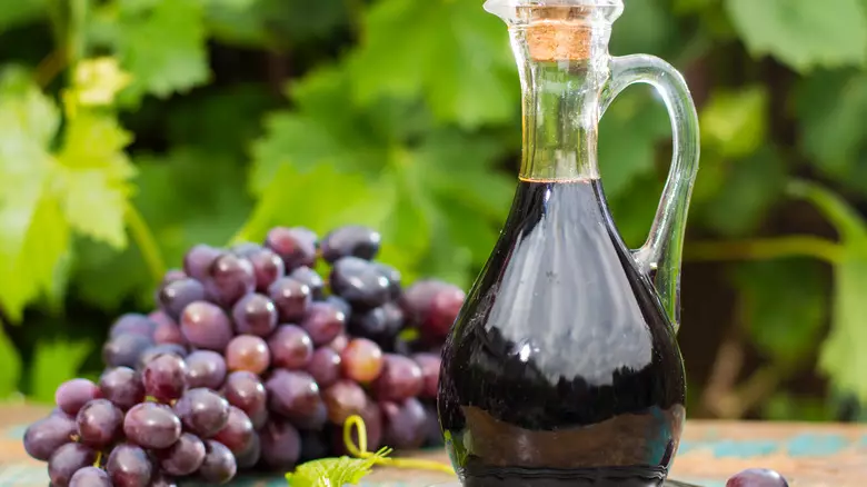 Botella de vinagre balsámico junto a una ristra de uvas frente a la vegetación