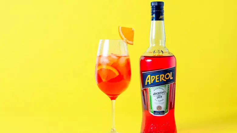 O sticlă de Aperol lângă un cocktail pe fond galben