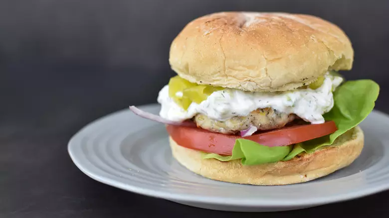 Un hamburger di pollo con lattuga, pomodori, salsa tzatziki, cipolle rosse e peperoni di banana sottaceto su un panino su un piatto bianco