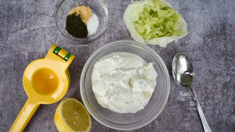 Uno spremiagrumi tenuto in mano accanto a un limone grattugiato, una ciotola di spezie, una ciotola di yogurt greco, alcuni cetrioli grattugiati e un cucchiaio