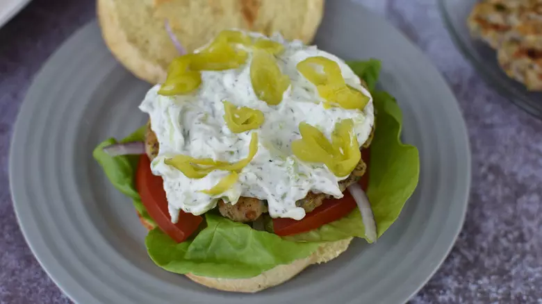 Atvērts vistas burgers ar salātiem, tomātiem, tzatziki, sarkanajiem sīpoliem un banānu pipariem uz zila šķīvja