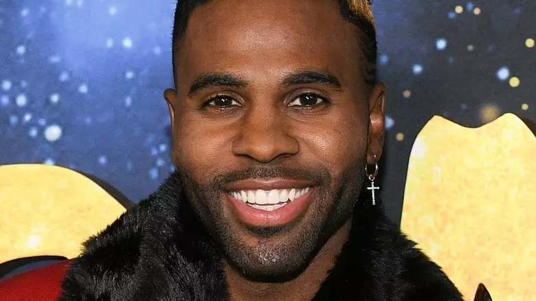 Ο Jason Derulo χαμογελά με σκουλαρίκι σταυρό και γούνινο μπουφάν