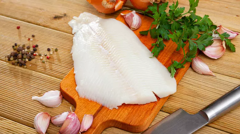 Fileo de kruda halibut-fiŝo sur ligna surfaco kun ajlo kaj verduloj