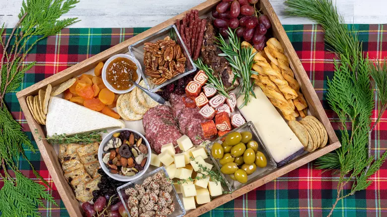 charcuterie board ndi tchizi, nyama, crackers, zipatso zouma, ma pecans okazinga, ma amondi otsekemera, ndi zina zowonjezera pa tebulo lobiriwira ndi lofiira la tartan ndi masamba atsopano.