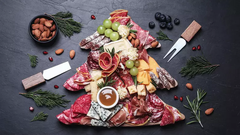 Mitengo ya Khrisimasi yopangidwa ndi charcuterie board yokhala ndi mitundu ingapo ya nyama zochiritsidwa ndi tchizi, zipatso zatsopano, ndi mtedza pamasamba akuda ndi masamba atsopano.