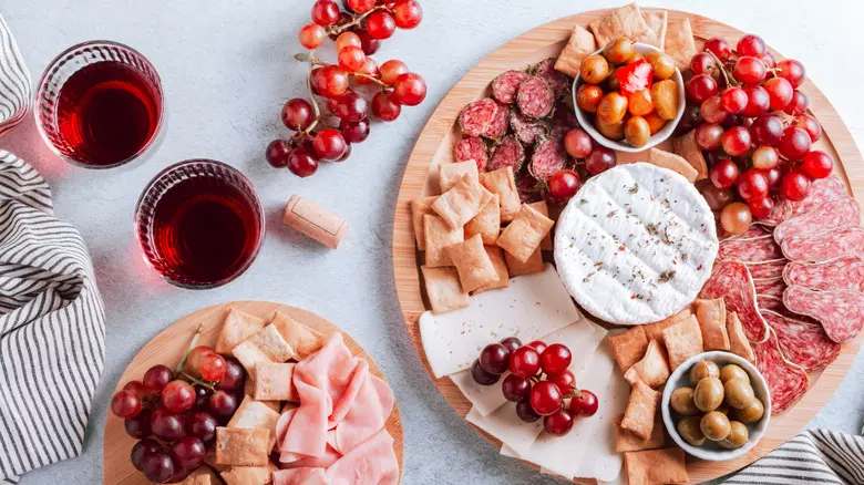 matabwa a charcuterie board ndi nyama zochiritsidwa, crackers, tchizi, azitona, ndi mphesa zofiira pa tebulo loyera ndi magalasi awiri a vinyo wofiira