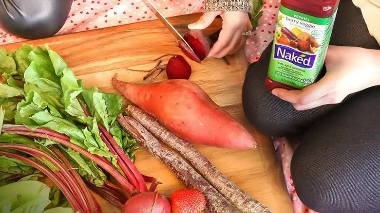 Mga utanon sa usa ka cutting board sa usa ka piknik, ug mga kamot nga nagkupot sa usa ka botelya sa berry veggie juice