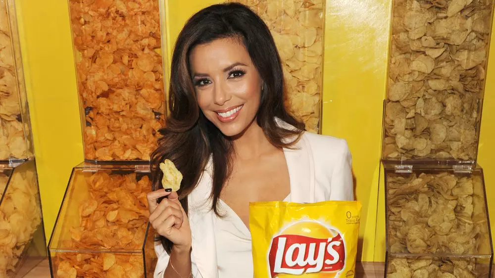 Eva Longoria dengan jaket dan kemeja putih memegang keripik kentang dan tas keripik kentang Lay